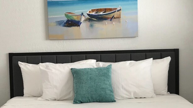 Uma cama com almofadas brancas e azul-turquesa, um cobertor vermelho e um quadro de barcos numa praia acima.