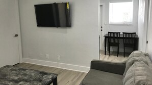 Uma sala de estar com televisão de tela plana na parede, um sofá cinzento, uma mesinha e um espaço de jantar com mesa e cadeiras.