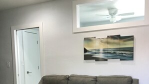 Sala de estar com sofá cinza, uma mesinha e um ventilador de teto. Há uma porta à esquerda e uma janela acima do sofá.