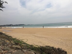 Uma praia arenosa com algumas pessoas caminhando, um trecho de costa rochosa e uma vista distante do oceano.