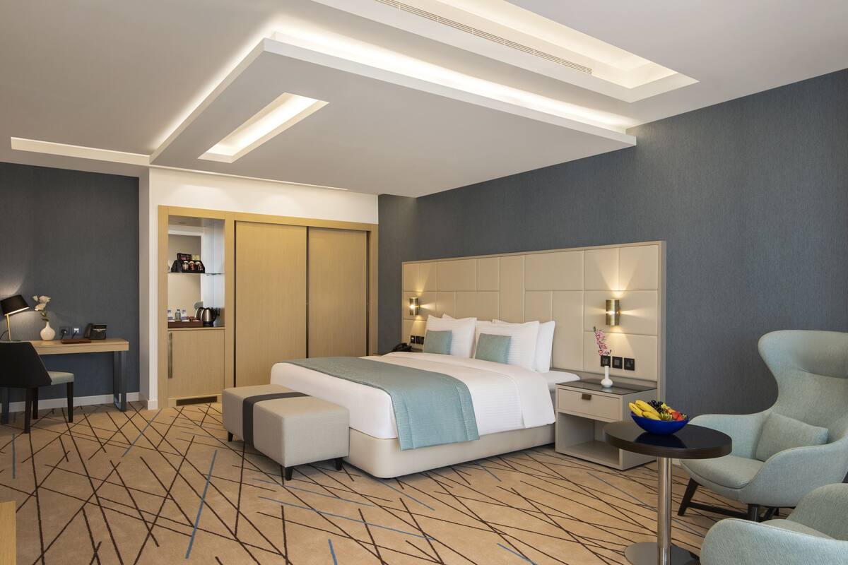 Quarto de hotel moderno com uma cama grande, uma escrivaninha, uma cadeira e uma pequena mesa com uma cesta de frutas.