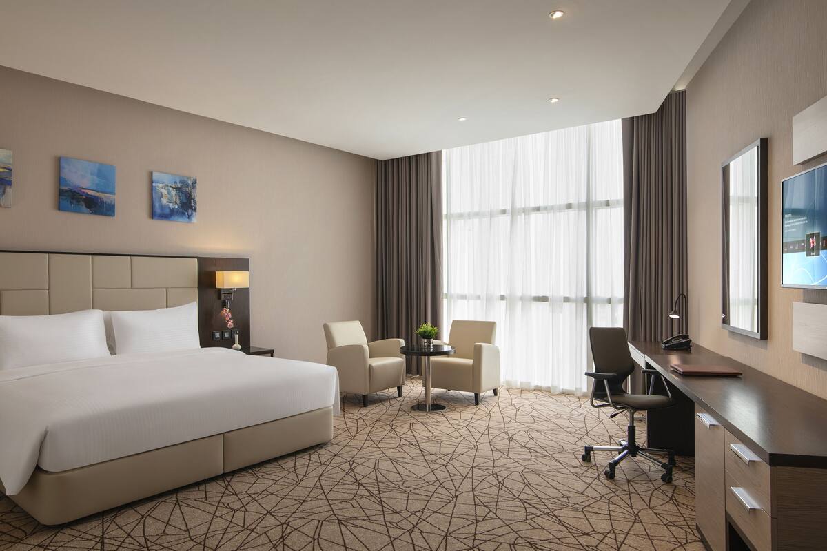 Quarto de hotel com uma cama grande, duas poltronas, uma escrivaninha e uma televisão.
