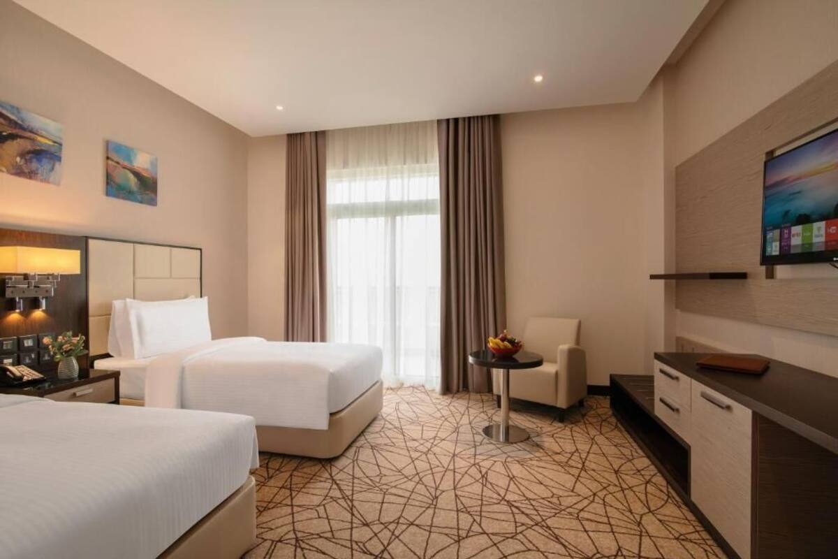 Quarto de hotel com duas camas, uma escrivaninha, uma cadeira, televisão e janela com cortinas.