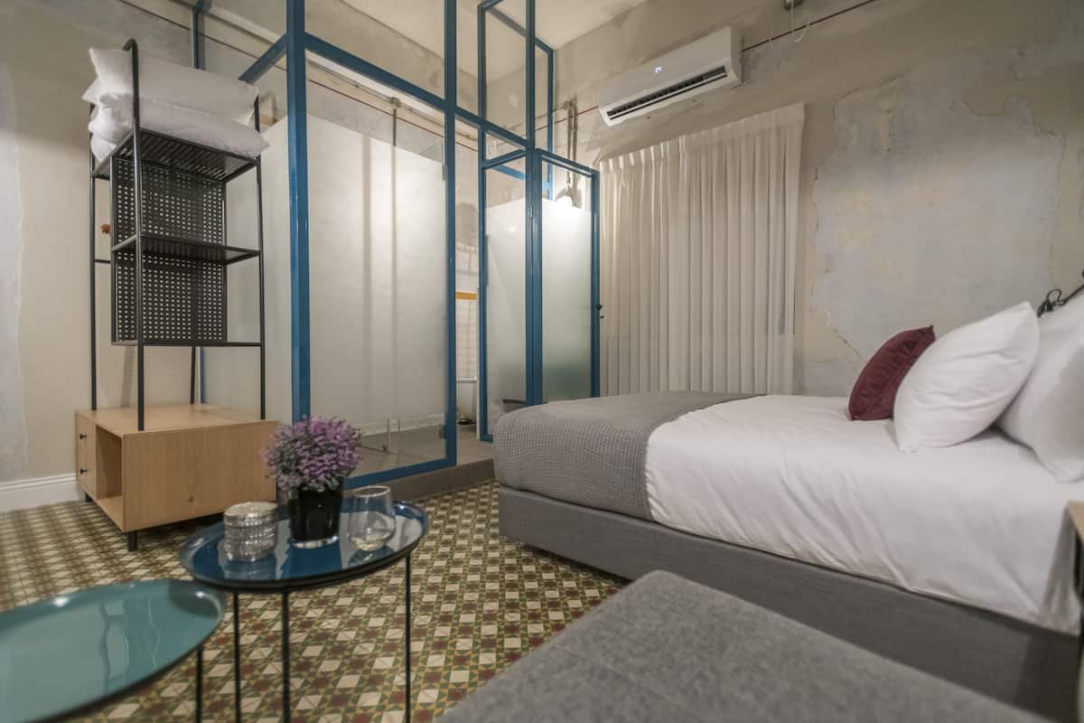 Quarto de hotel moderno com sofá cinzento, uma pequena mesa lateral de madeira, um espelho e um chão com padrão.