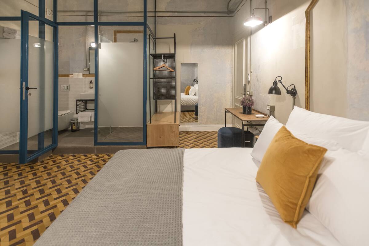 Quarto de hotel moderno com uma cama, uma mesa de escritório e um banheiro visível através de uma porta aberta.