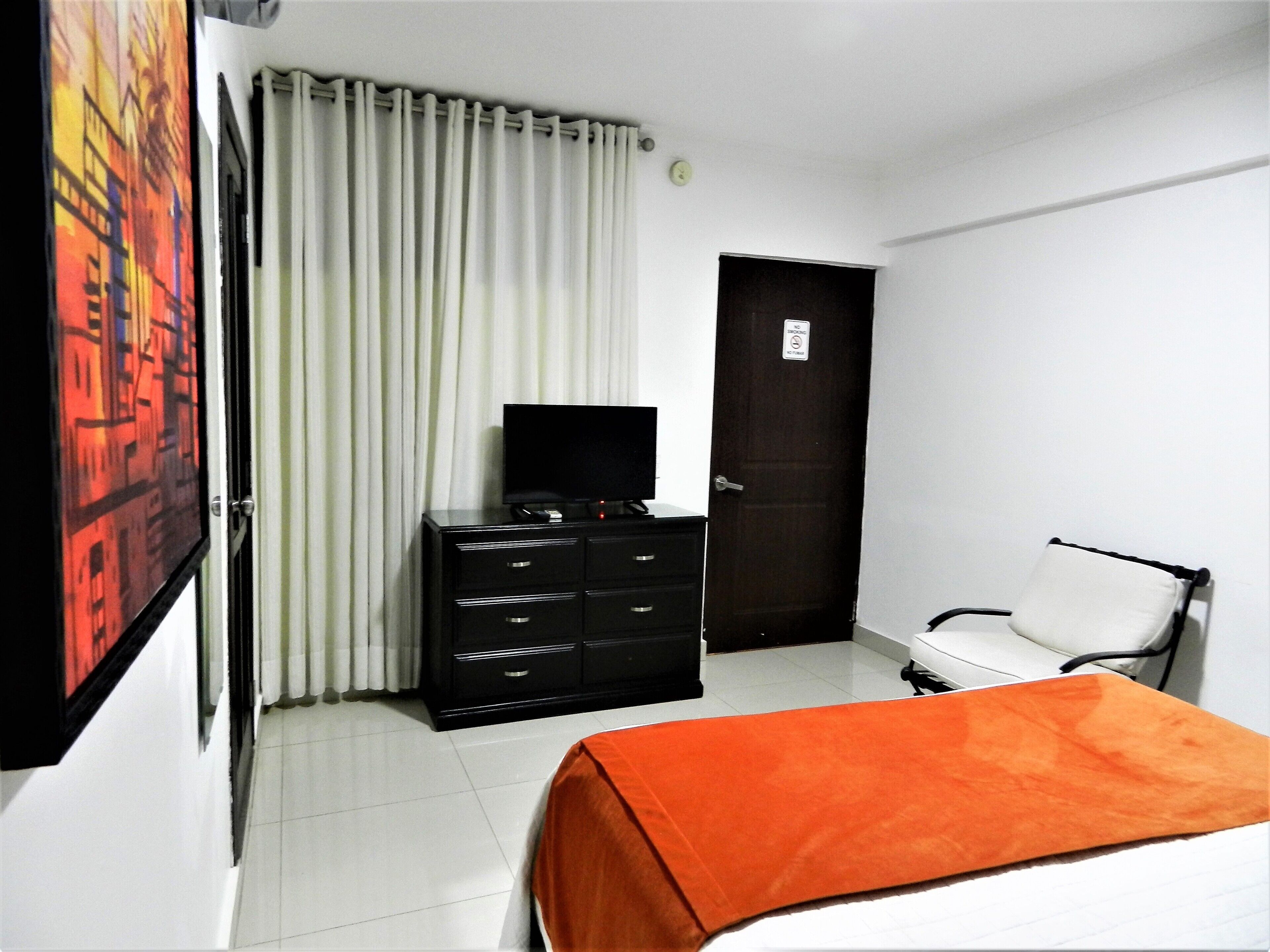 Quarto de hotel com uma cama, televisão em uma cômoda, uma cadeira, uma porta e uma cortina.