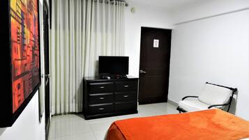 Quarto de hotel com uma cama, televisão em uma cômoda, uma cadeira, uma porta e uma cortina.