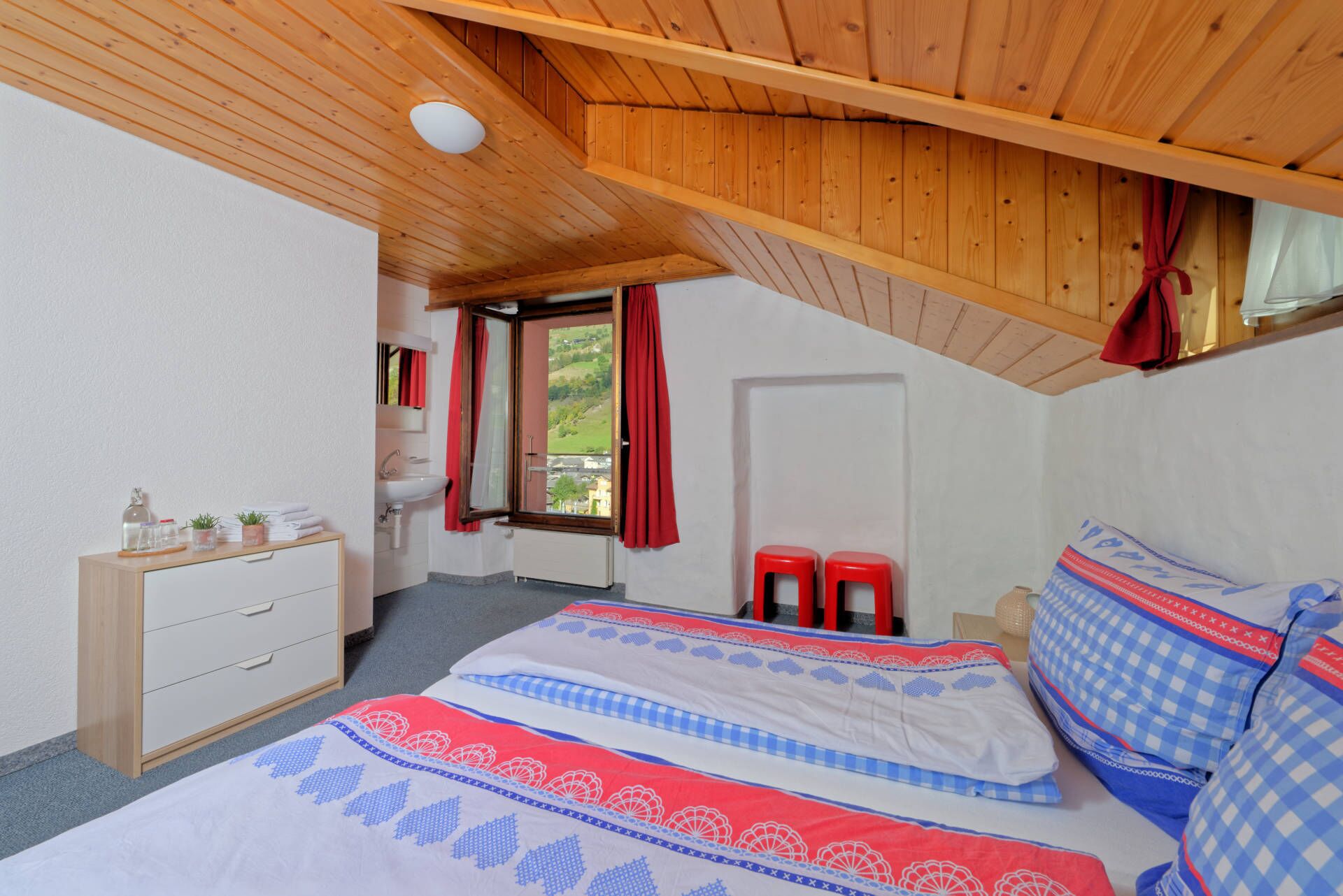 Quarto com teto de madeira, cama com roupa de cama estampada, mesa de cabeceira com luminária e janela com cortinas vermelhas.