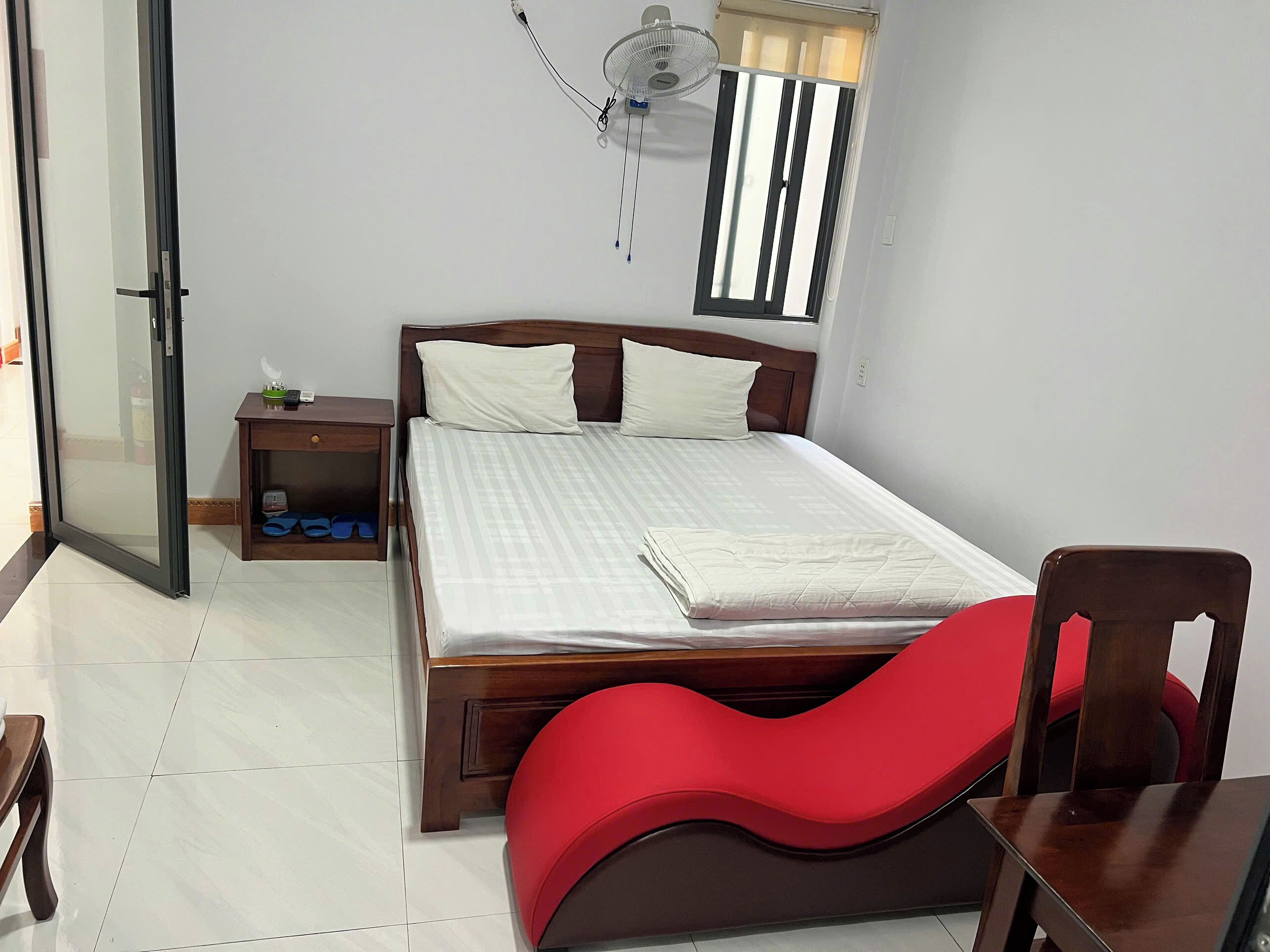 Quarto com cama, mesa de cabeceira, cadeira, um banco vermelho e um ventilador.