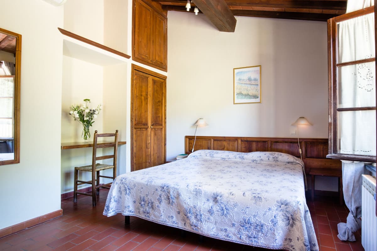 Quarto com cama de madeira, uma cadeira, uma mesa e janela com cortinas.
