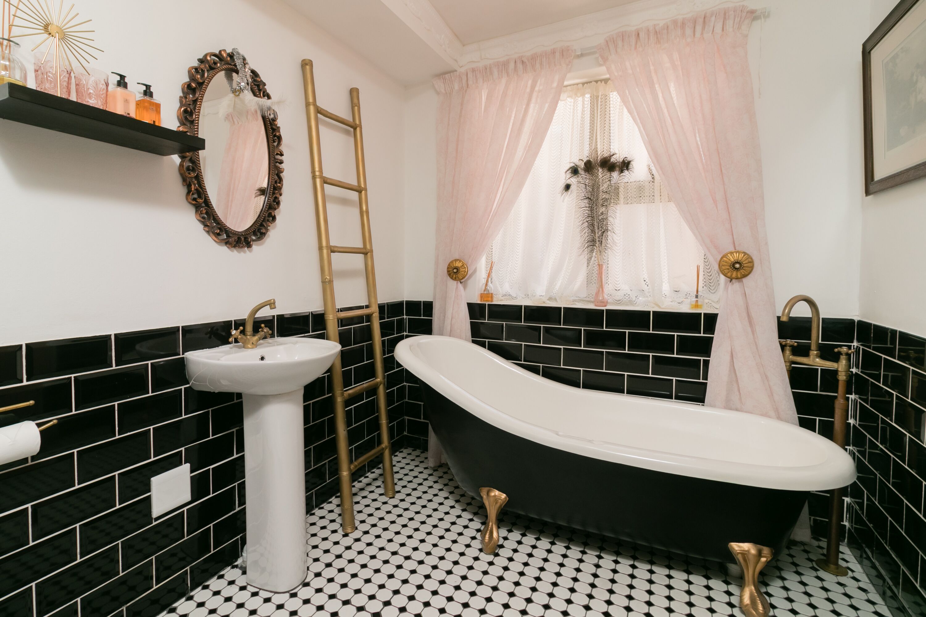 Um banheiro com piso xadrez preto e branco, pia branca de pedestal, banheira independente preta, escada dourada e janela com cortinas leves.