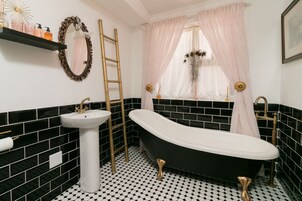 Um banheiro com piso xadrez preto e branco, pia branca de pedestal, banheira independente preta, escada dourada e janela com cortinas leves.