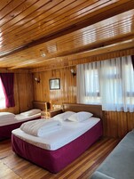 Quarto estilo cabana com painéis de madeira, duas camas com lençóis brancos, uma cabeceira de madeira, uma mesinha de cabeceira, um abajur e uma janela com cortinas leves.
