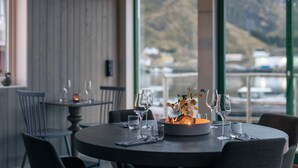 Mesa de jantar posta com copos e um arranjo central, com vista para barcos e montanhas através de amplas janelas.