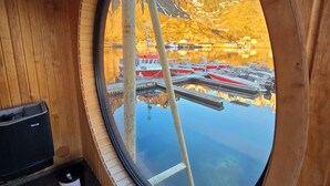 Uma cabana de madeira com janela circular que oferece vista para barcos e montanhas.