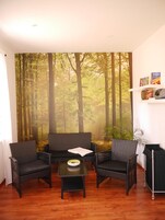 Um quarto aconchegante com um mural temático de floresta, duas cadeiras de vime, uma mesinha com uma tigela e uma estante de livros com livros.