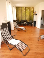 Quarto com piso de madeira, duas poltronas listradas em preto e branco, uma mesinha com uma tigela e guardanapos, uma estante de livros e vista para uma floresta através de uma grande janela.