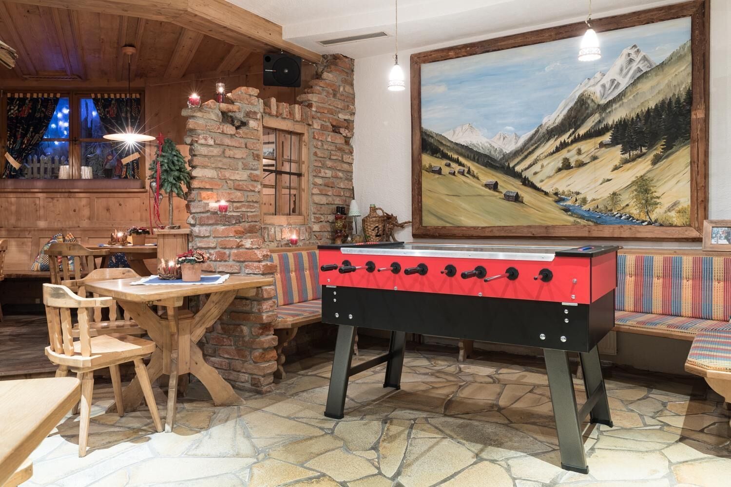 Um ambiente acolhedor com mesa de futebol, móveis de madeira, piso de pedra e uma grande pintura de paisagem de montanhas na parede.