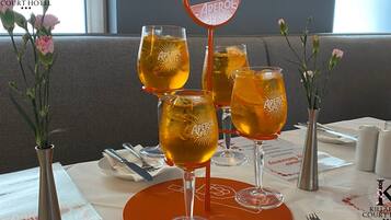 Mesa posta com quatro copos de Aperol Spritz, um vaso com flores rosa e uma placa com o nome do hotel.