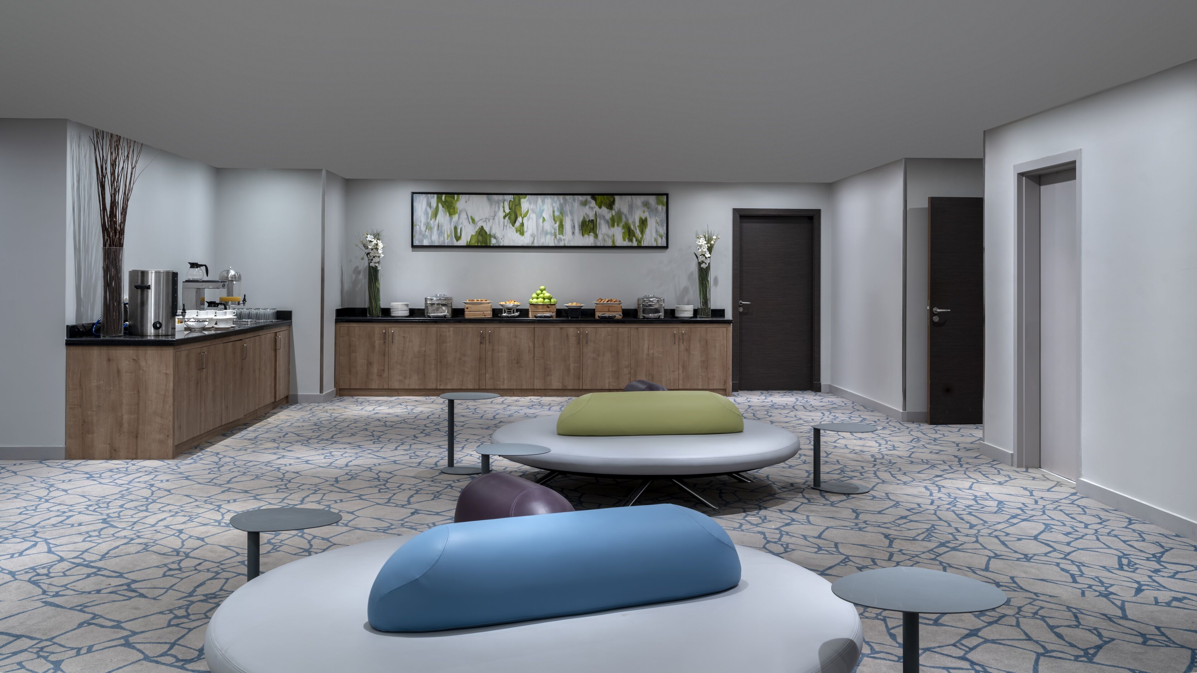 Um saguão de hotel moderno com uma área de estar central, um balcão de cozinha e uma grande pintura abstrata na parede.