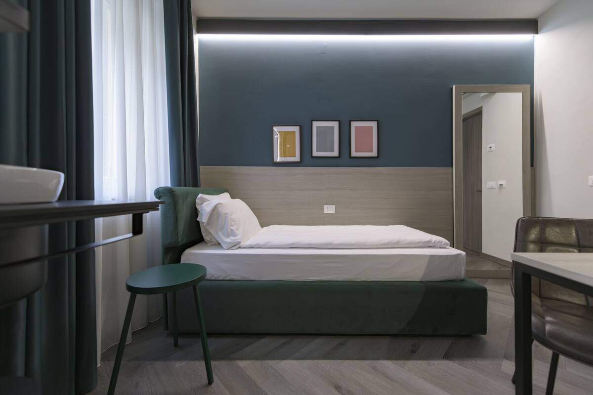 Quarto compacto com cama, um banquinho pequeno, uma mesa e um espelho.