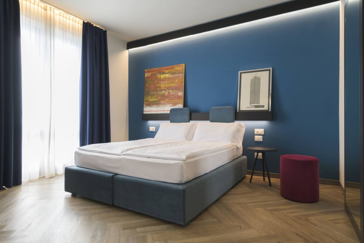 Quarto de hotel moderno com uma cama grande, uma pequena mesa de apoio, um banquinho vermelho e uma pintura com vista para a cidade na parede.
