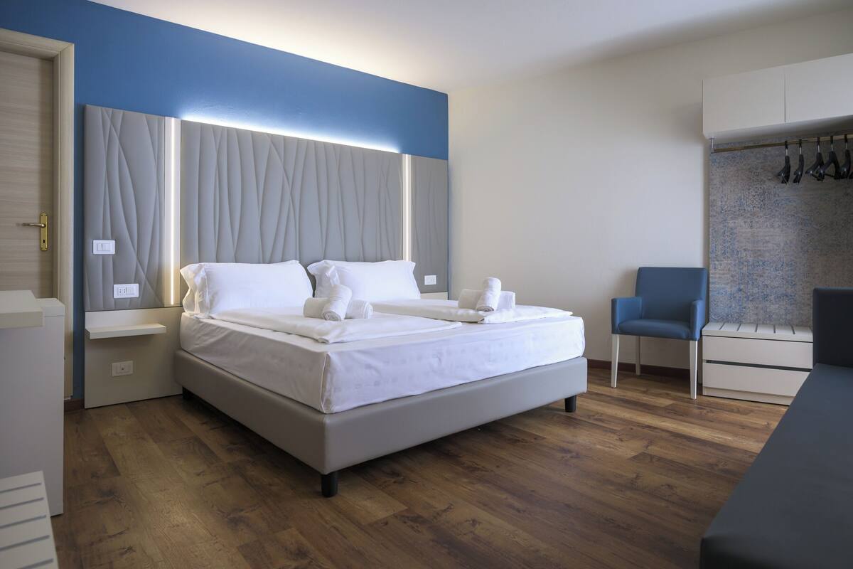 Quarto de hotel moderno com uma cama grande, mesas de cabeceira, uma cadeira e um guarda-roupa.
