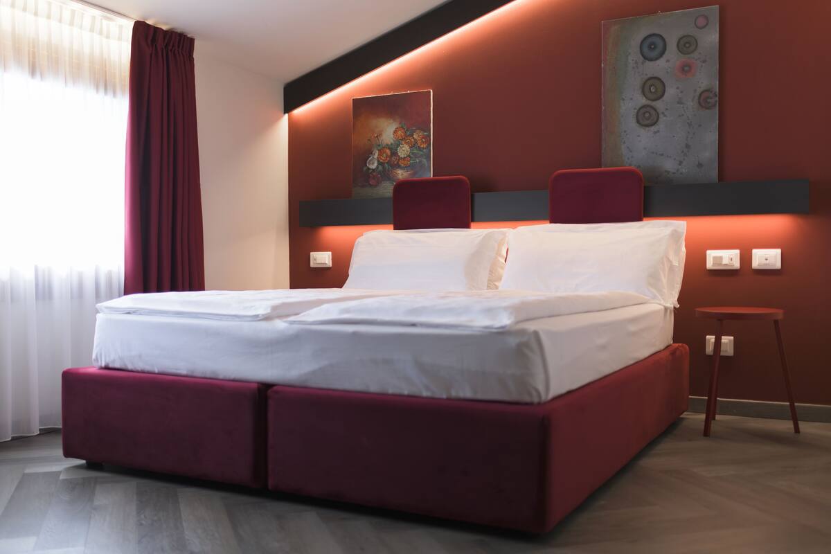 Quarto de hotel moderno com cama grande, cabeceira estofada em vermelho e uma pintura na parede.