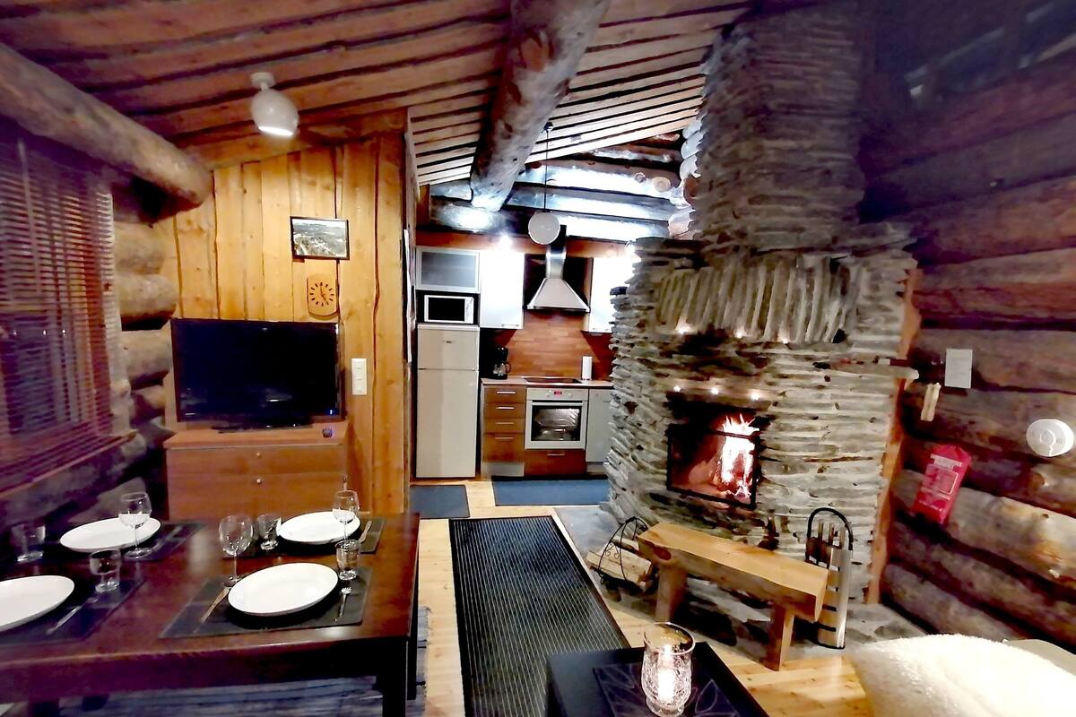 Interior rústico de uma cabana com  área de jantar de madeira, lareira de pedra, televisão e uma kitchenette.