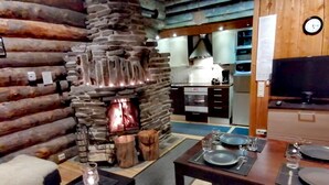 Uma área de refeições acolhedora, com parede de pedra, toras de madeira e uma cozinha moderna ao fundo.