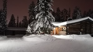 Uma cabana de toras com telhado coberto de neve e entrada iluminada, rodeada por neve e árvores.