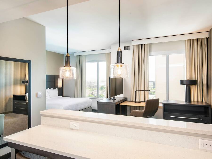 Quarto de hotel com cama, escrivaninha, cadeira e janelas amplas.