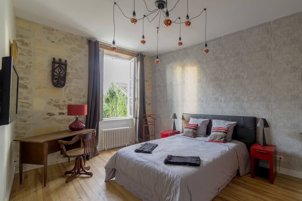 Um quarto com cama, escrivaninha, cadeira, luminária vermelha e lustre.