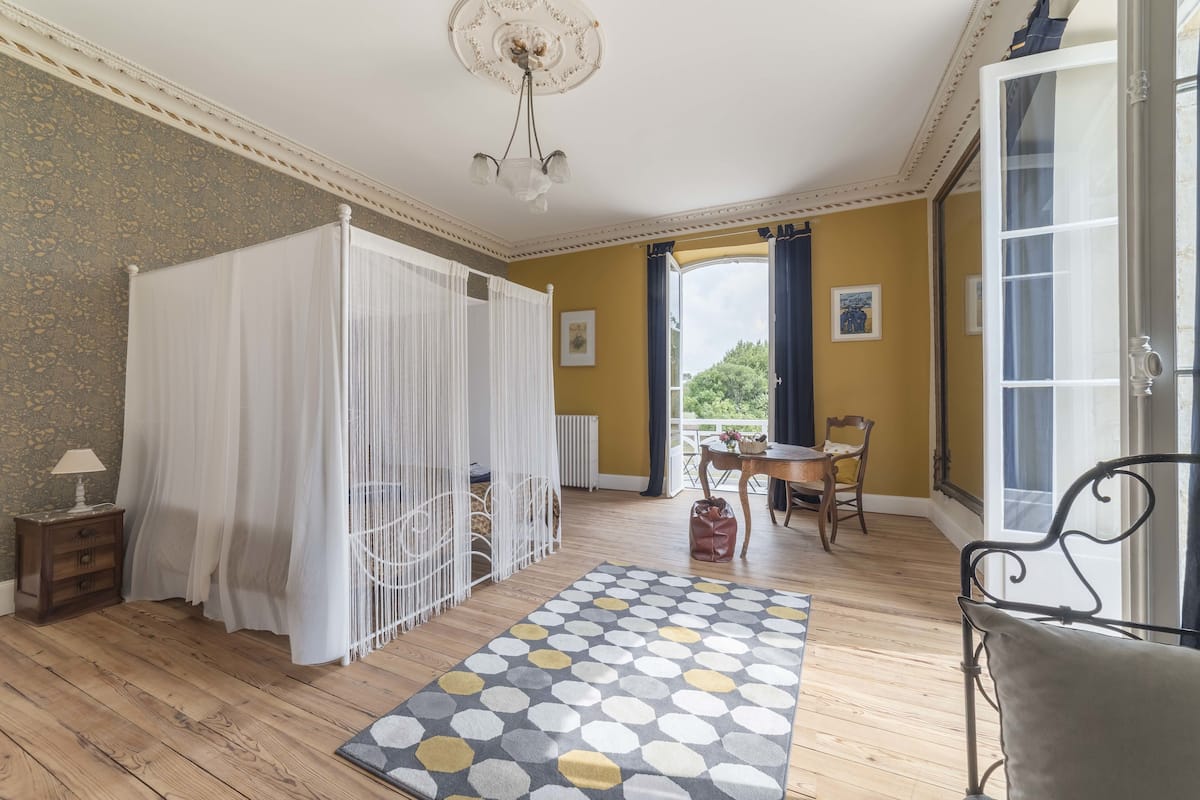 Quarto com cama de dossel, piso de madeira, tapete estampado, mesa de jantar com cadeiras e vista para vegetação pela janela.