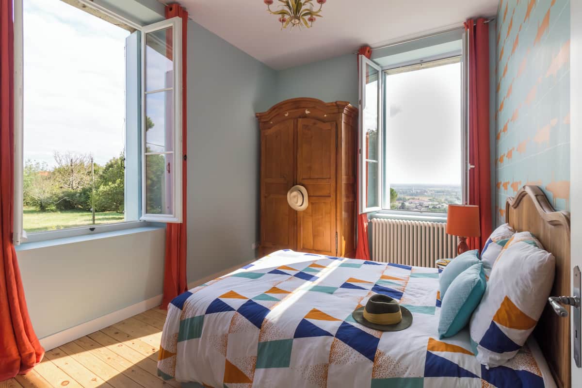 Quarto com uma janela grande, guarda-roupa de madeira, cama com colcha e vista para o exterior.