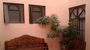 Quarto com sofá de madeira, uma poltrona combinando e uma mesa pequena. Há duas janelas com persianas e um vaso de planta.