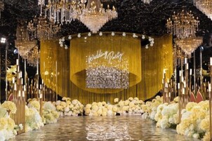 Palco com fundo dourado e um letreiro 'Wedding Party', rodeado por arranjos florais brancos.