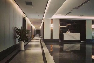 Um hall de hotel com balcão de recepção, planta em vaso e um corredor longo.