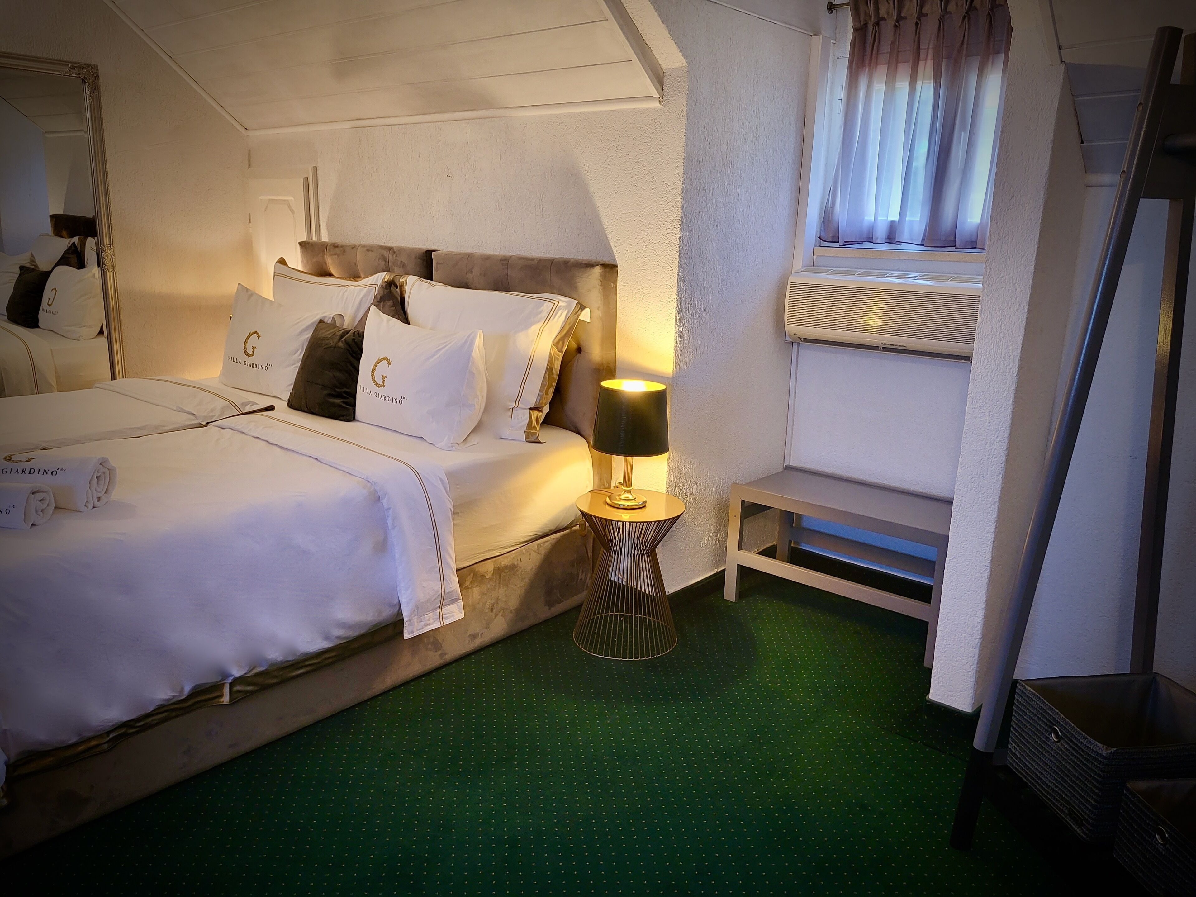 Quarto de hotel com cama, almofadas, abajur, mesinha de cabeceira e janela com cortinas.