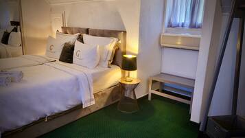 Quarto de hotel com cama, almofadas, abajur, mesinha de cabeceira e janela com cortinas.