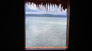Vista pela janela com telhado de palha, com vista para águas calmas e montanhas ao longe.
