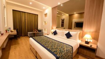 Quarto de hotel com uma cama grande, mesa de cabeceira com abajur, escrivaninha com cadeira, um sofá pequeno e uma janela ampla com cortinas.