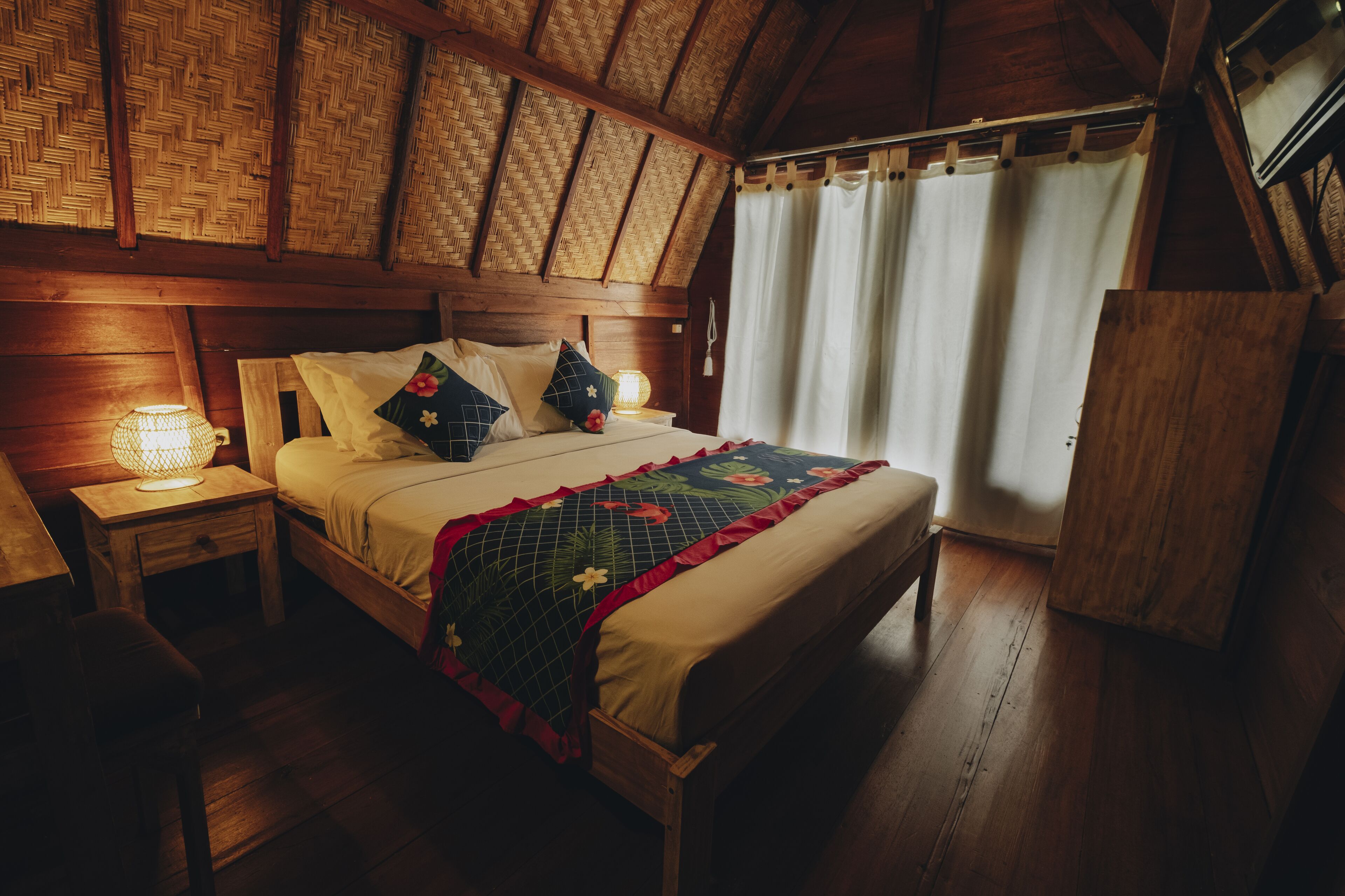 Uma cama de madeira com colcha floral, uma mesa de cabeceira com abajur e uma janela com cortinas.