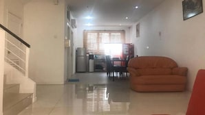 Uma sala ampla com um sofá vermelho, uma mesa de jantar com cadeiras, uma cozinha com geladeira e uma escada à esquerda.