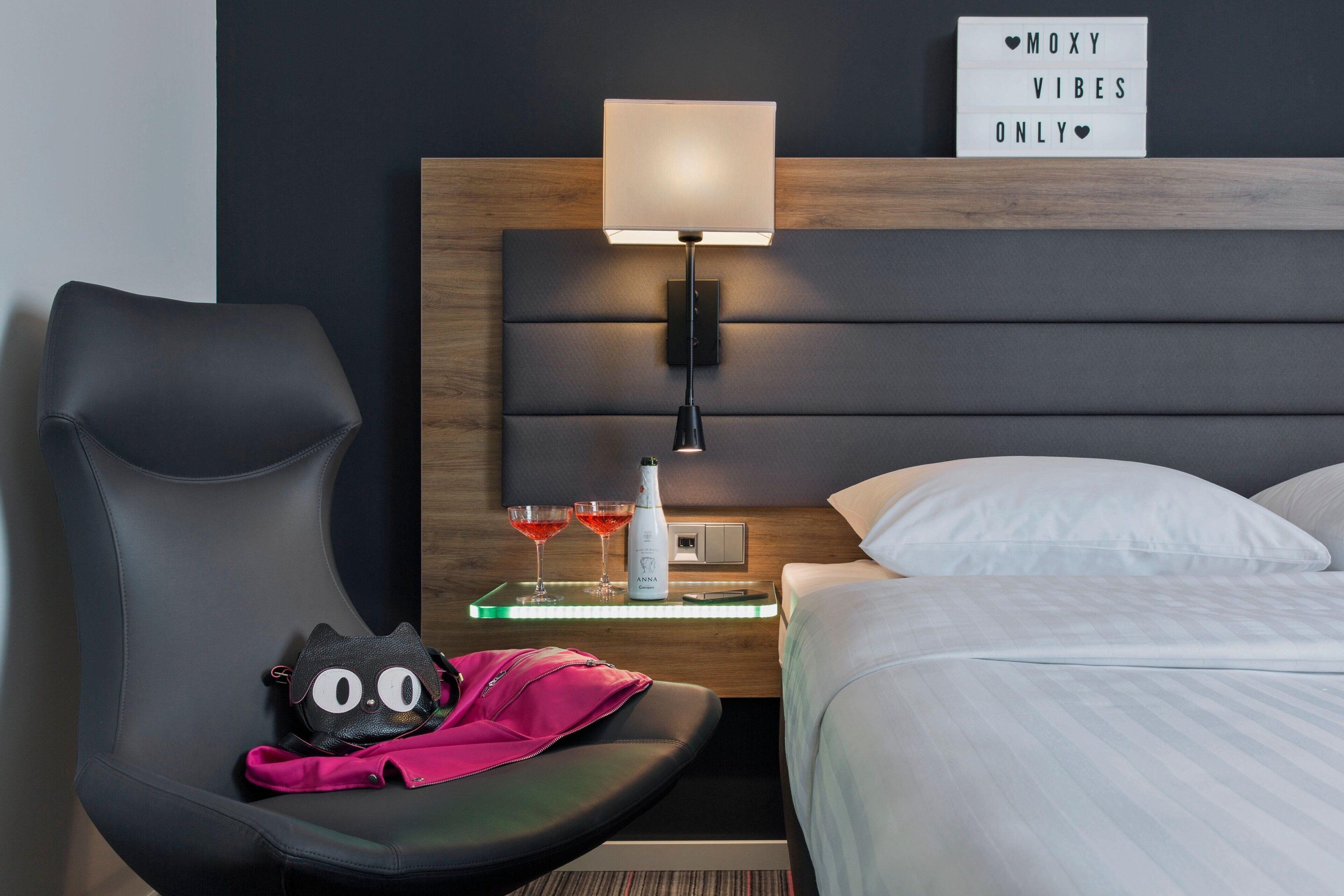 Quarto de hotel com design moderno, incluindo uma cama, uma cadeira com travesseiro em formato de gato, uma mesa de cabeceira com abajur e uma placa com a inscrição 'MOXY VIBES ONLY'.