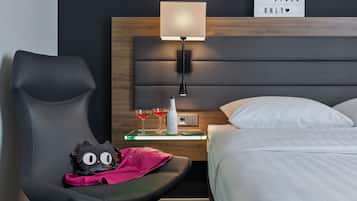 Quarto de hotel com design moderno, incluindo uma cama, uma cadeira com travesseiro em formato de gato, uma mesa de cabeceira com abajur e uma placa com a inscrição 'MOXY VIBES ONLY'.