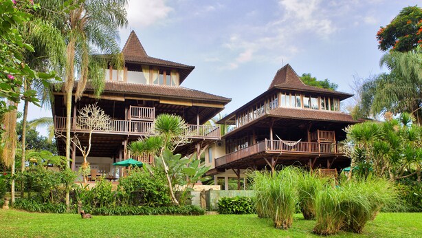 Um edifício de dois andares com varandas, rodeado por vegetação exuberante e um gramado bem cuidado.