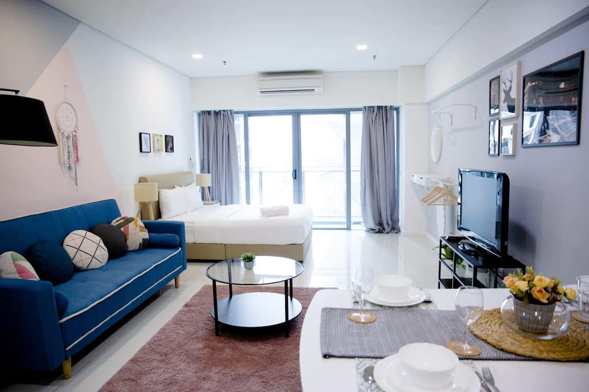 Quarto de hotel moderno com sofá azul, cama com roupa de cama branca, mesa de centro de vidro, área de jantar com mesa posta para dois e televisão sobre um móvel.