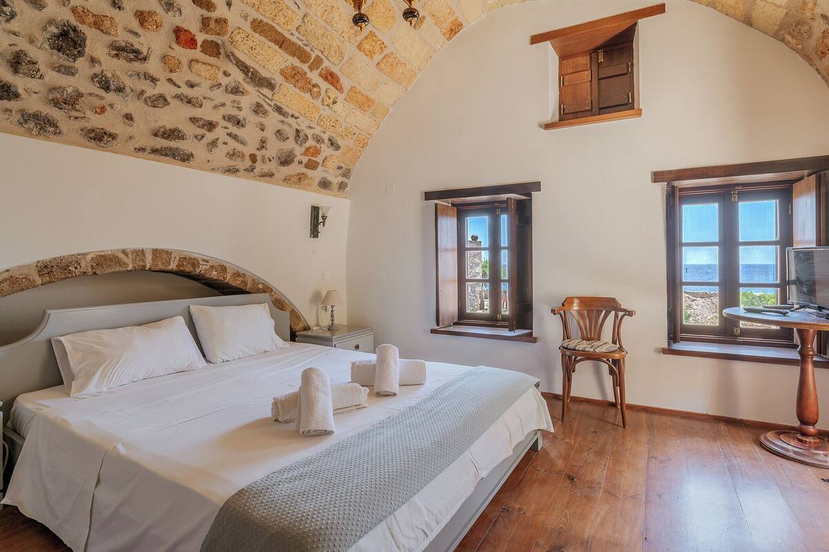 Quarto com teto de pedra, cama com lençóis brancos, duas janelas com vista e uma cadeira de madeira.