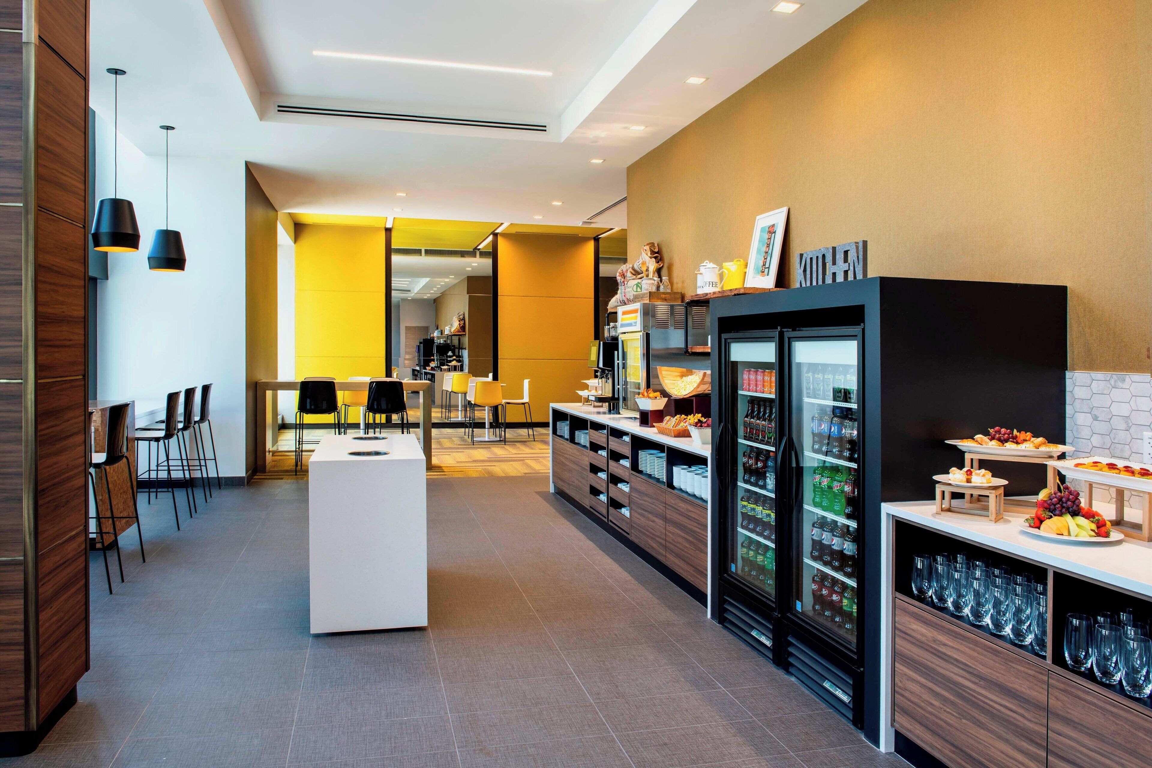 Um café moderno com um balcão central branco, área de estar com cadeiras, cozinha com refrigerador cheio de bebidas e um buffet com frutas e doces.